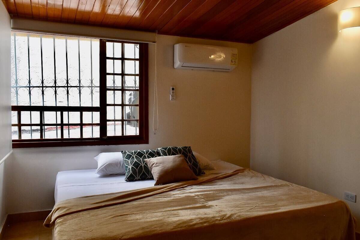 Quarto com cama, janela com grades, ar condicionado e luminária fixada na parede.