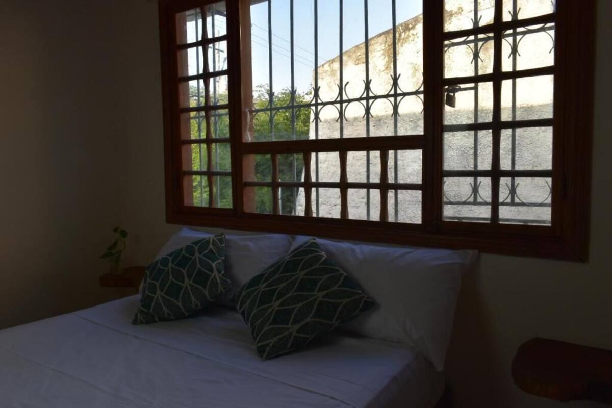 Quarto com cama, duas almofadas estampadas, mesa de cabeceira com uma planta e janela com grades de metal.