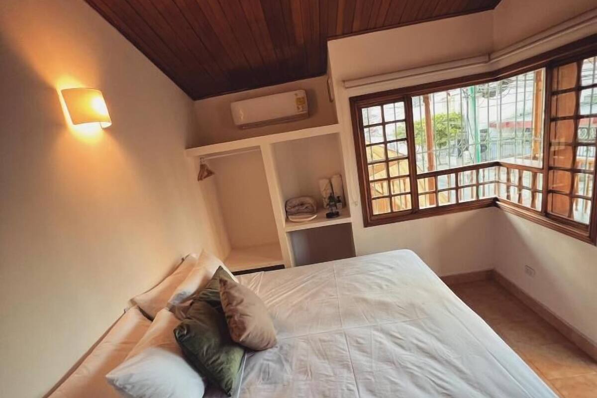 Quarto com teto de madeira, cama com almofadas, mesa de cabeceira e janela com vista para o exterior.