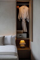 Quarto de hotel com cama bem arrumada, luminária de cabeceira, roupão pendurado no armário e uma pequena mesa com uma cesta.