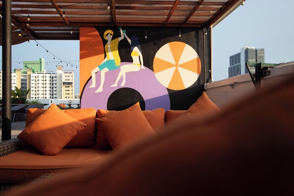 Espaço de estar no terraço com almofadas laranja e um mural colorido.