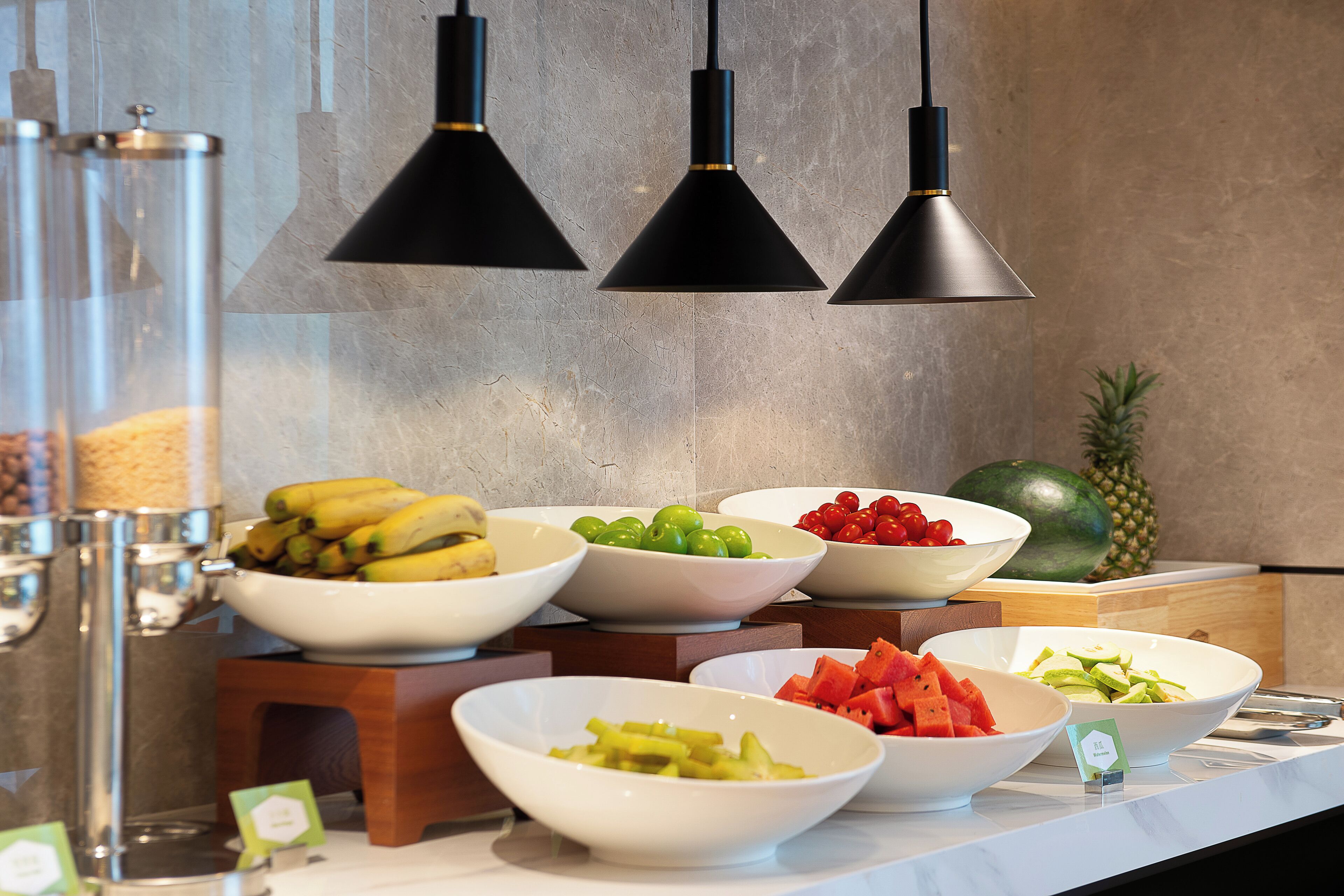 Buffet com diversas frutas e legumes, incluindo bananas, melancia e maçãs verdes, dispostos sobre um balcão com detalhes em mármore e iluminação pendente.