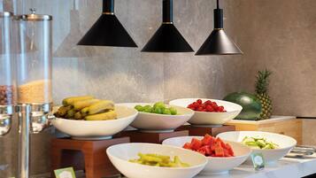 Buffet com diversas frutas e legumes, incluindo bananas, melancia e maçãs verdes, dispostos sobre um balcão com detalhes em mármore e iluminação pendente.