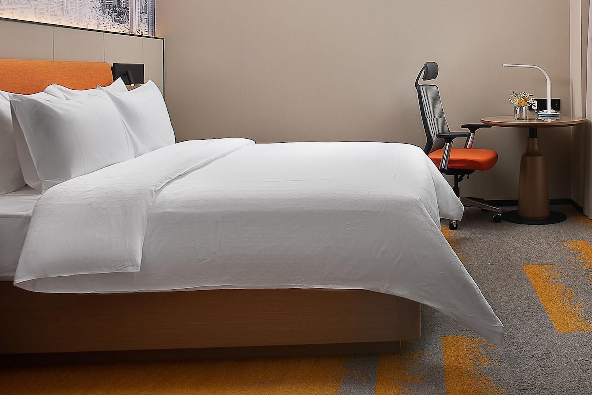 Quarto de hotel com uma cama, uma mesinha de cabeceira, uma poltrona laranja, uma pequena mesa com um vaso e uma luminária na parede.