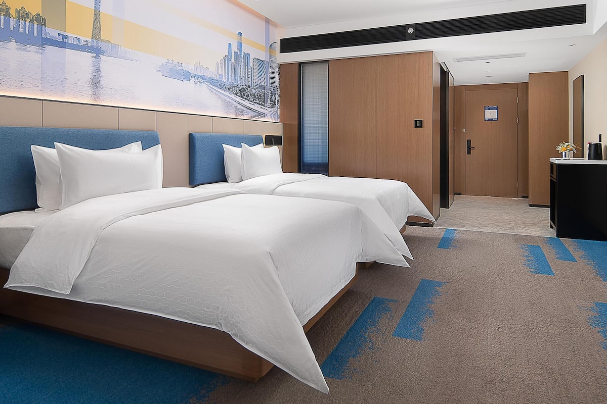 Quarto de hotel com duas camas, um mural grande na parede, uma escrivaninha com um vaso e uma porta que dá para outro cômodo.