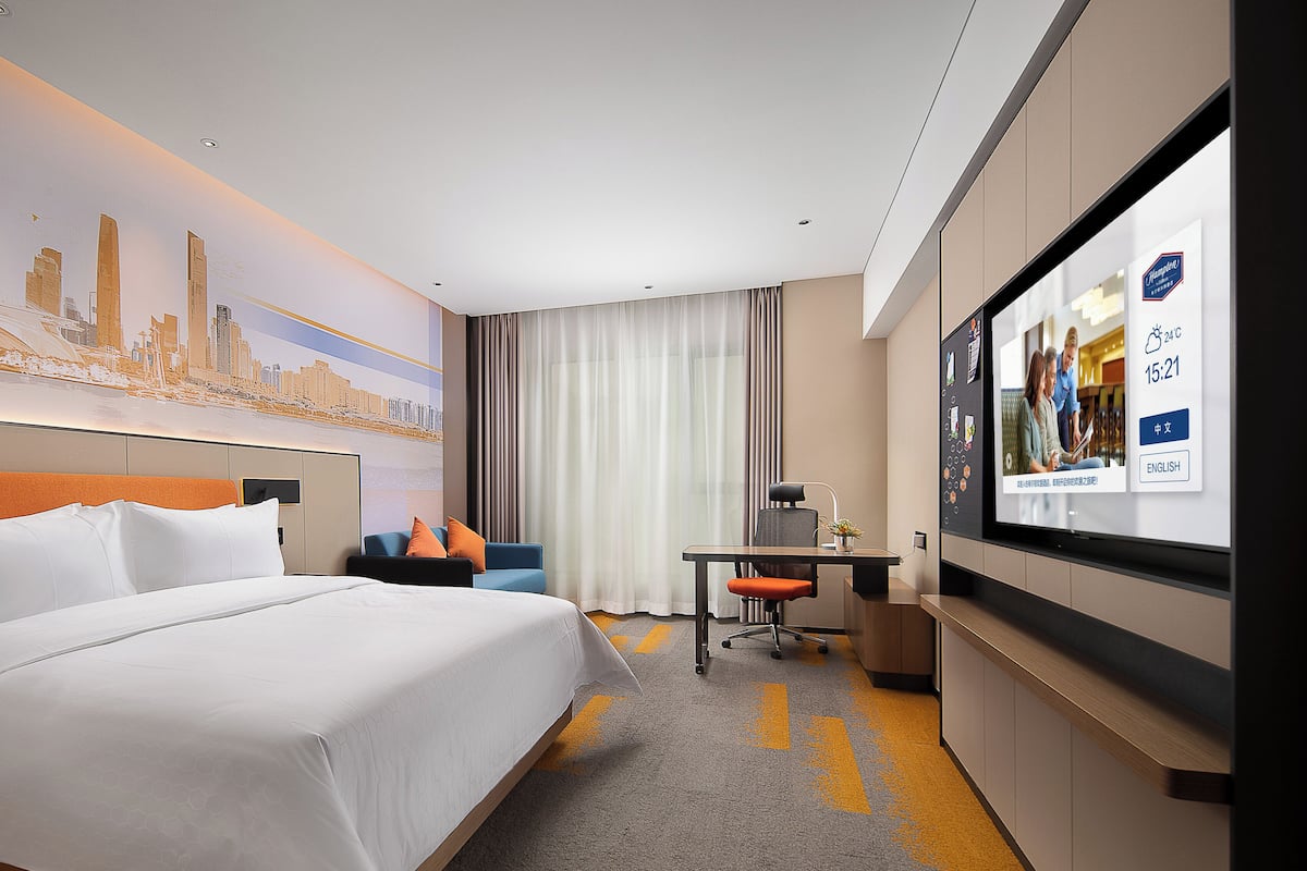 Quarto de hotel com cama, escrivaninha, cadeira, televisão e um mural com vista urbana na parede.
