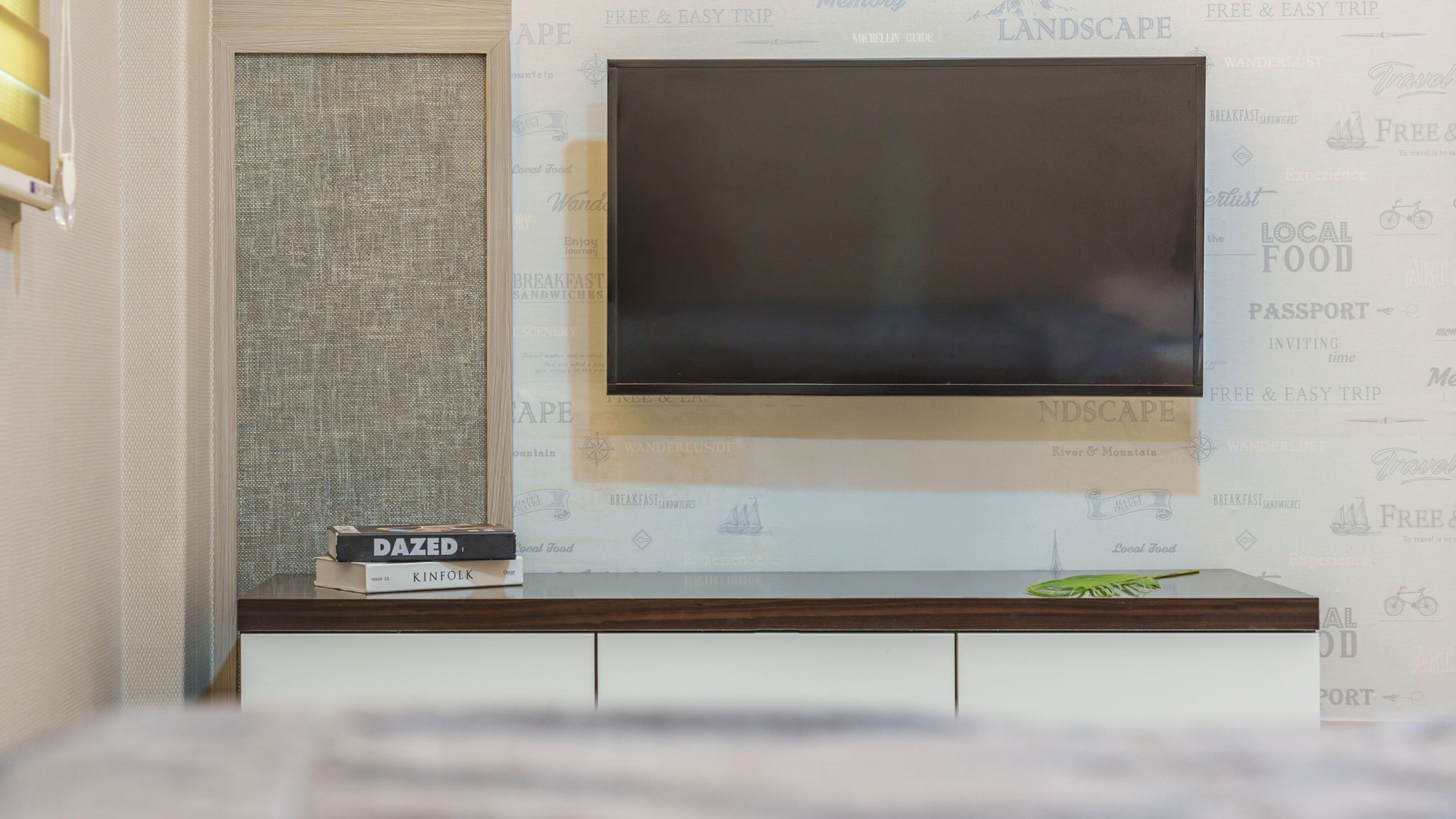 Uma sala de estar moderna com uma TV de tela plana na parede, uma mesinha de centro com uma revista DAZED e uma parede decorativa com papel de parede de tema de viagens.