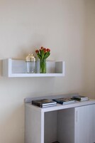 Uma prateleira branca com um vaso de flores vermelhas e um recipiente de vidro transparente. Abaixo, um armário branco com livros e uma pequena mesa são visíveis.