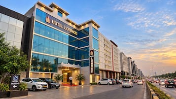 Um hotel moderno com fachada de vidro, entrada com tapete vermelho e vários carros estacionados.