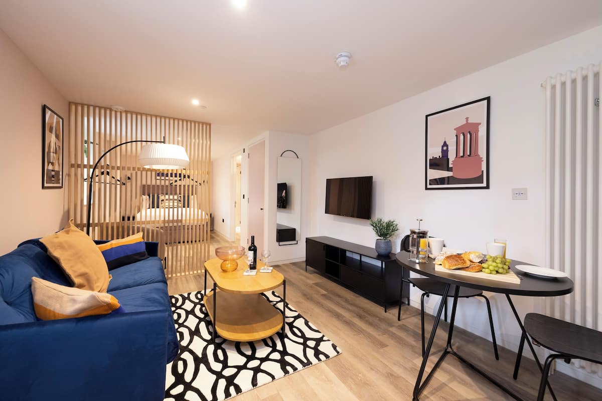 Uma sala de estar moderna com um sofá azul, uma mesa de jantar de madeira com um vaso e uma TV preta na parede.