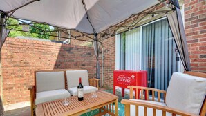 Um pátio externo com mesa de madeira, duas cadeiras estofadas, uma garrafa de vinho e um cooler vermelho Coca-Cola.