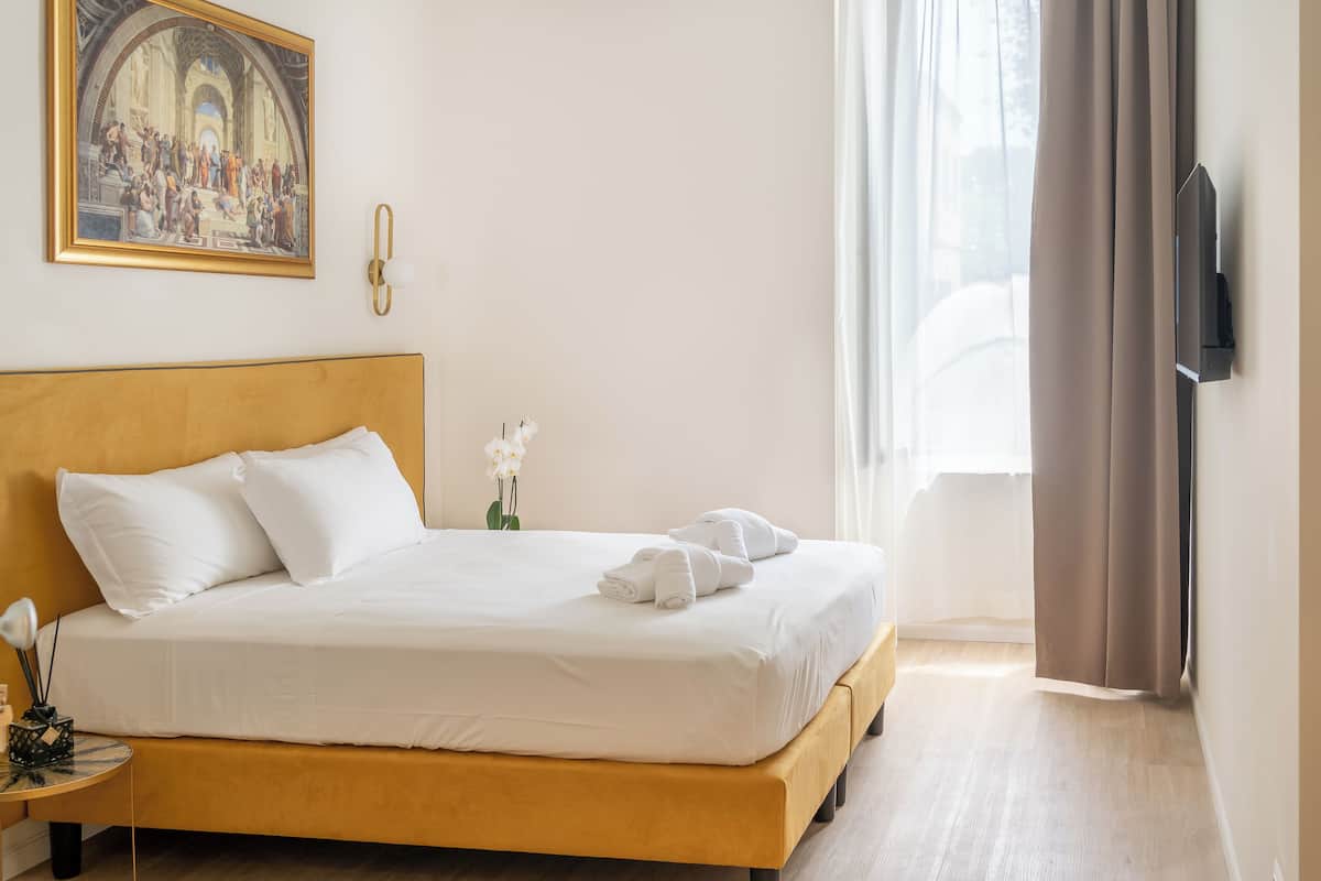 Quarto de hotel com cama, quadro na parede, televisão e janela com cortinas.