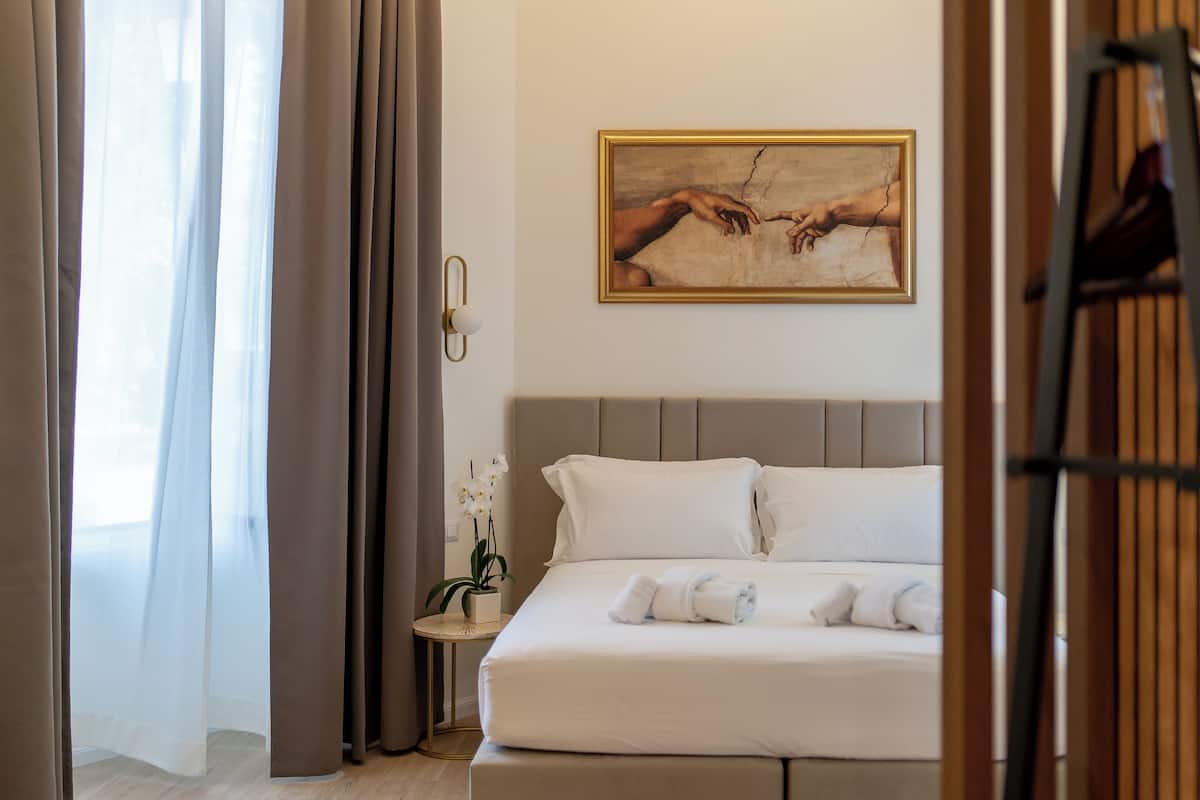 Quarto de hotel com cama, quadro na parede, mesinha com uma planta e janela com cortinas.