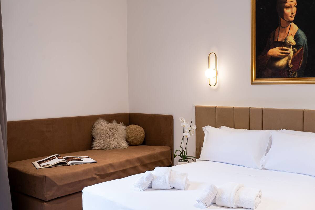 Quarto de hotel com uma cama, um sofá marrom, um quadro na parede e uma orquídea em vaso.