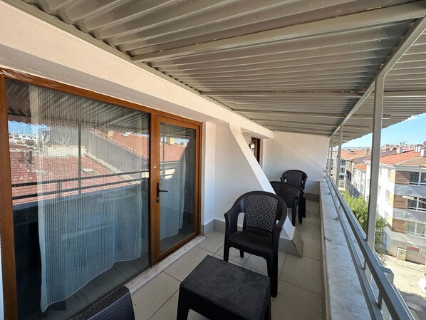 Varanda com porta de correr de vidro, duas cadeiras de vime, uma mesa pequena e vista para edifícios.