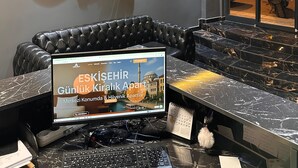 Uma área de recepção moderna com um sofá de couro, um computador exibindo um anúncio de imóvel e uma motocicleta ao fundo.