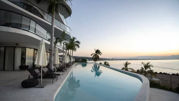 Piscina moderna de hotel com espreguiçadeiras e guarda-sóis, com vista para uma praia e oceano ao pôr do sol.