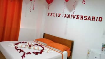 Um quarto com uma cama, roupa de cama laranja, uma parede branca com a inscrição 'FELIZ ANIVERSARIO' e balões em formato de coração vermelhos.