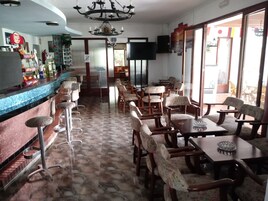 Um restaurante com área de bar, diversas opções de assento incluindo banquetas e mesas, um lustre e uma televisão na parede.