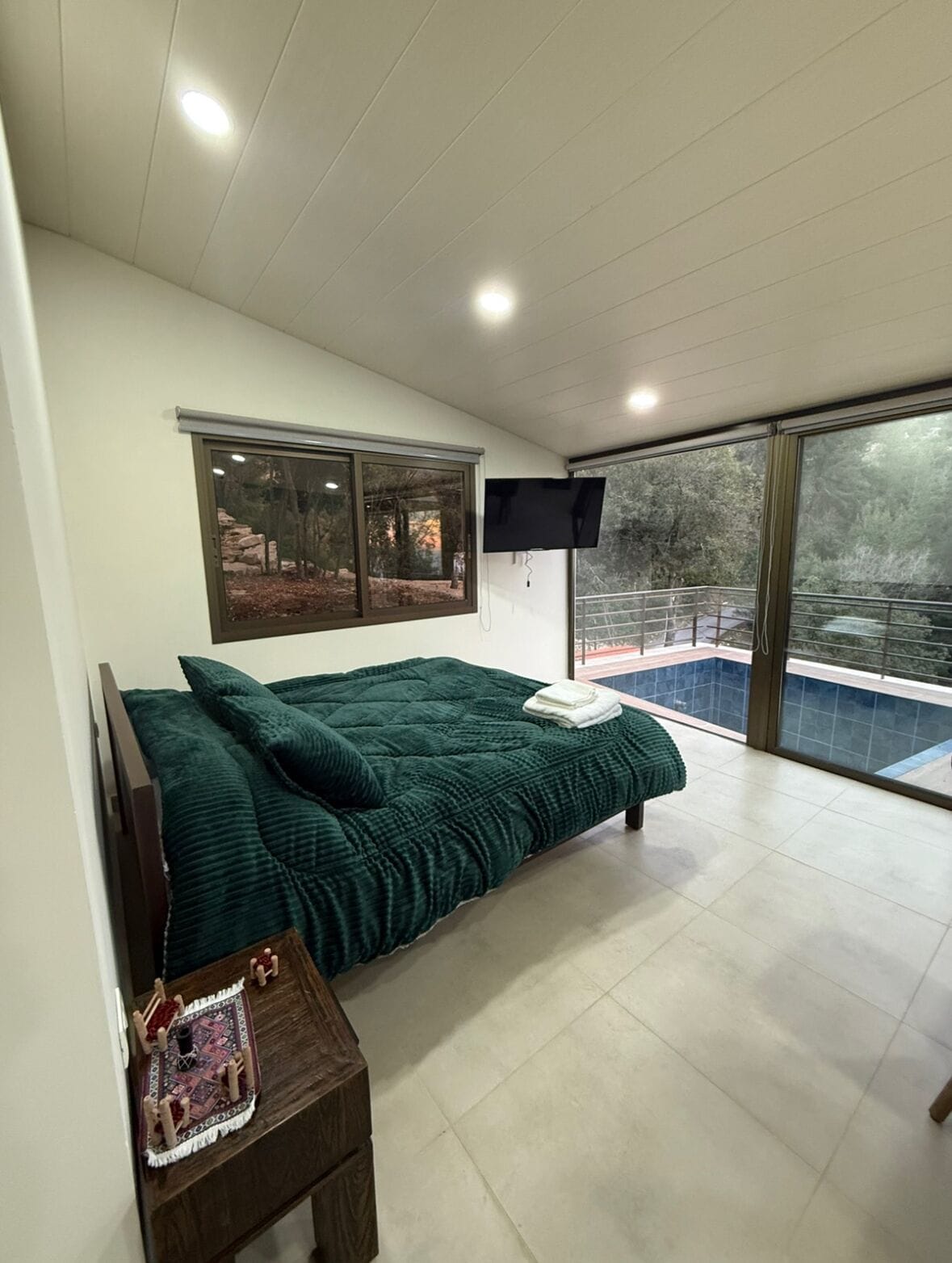 Quarto com uma cama grande, TV fixada na parede e vista para uma piscina através de janelas amplas.