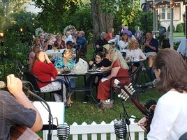 Um grupo ao ar livre com pessoas sentadas em mesas, algumas com instrumentos musicais, em um jardim.