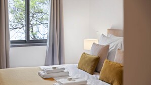 Uma cama bem arrumada com roupa de cama branca e cabeceira de madeira, uma luminária de cabeceira e uma janela com cortinas.