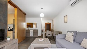 Uma sala de estar moderna com um sofá cinzento, mesa de centro de madeira e uma TV sobre um armário de madeira. Há uma área de jantar com mesa e cadeiras, e uma cozinha ao fundo.