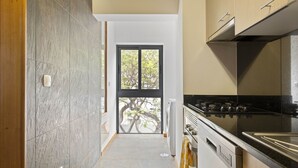 Uma cozinha moderna com fogão, forno e máquina de lavar loiça. Uma janela com vista para árvores é visível.