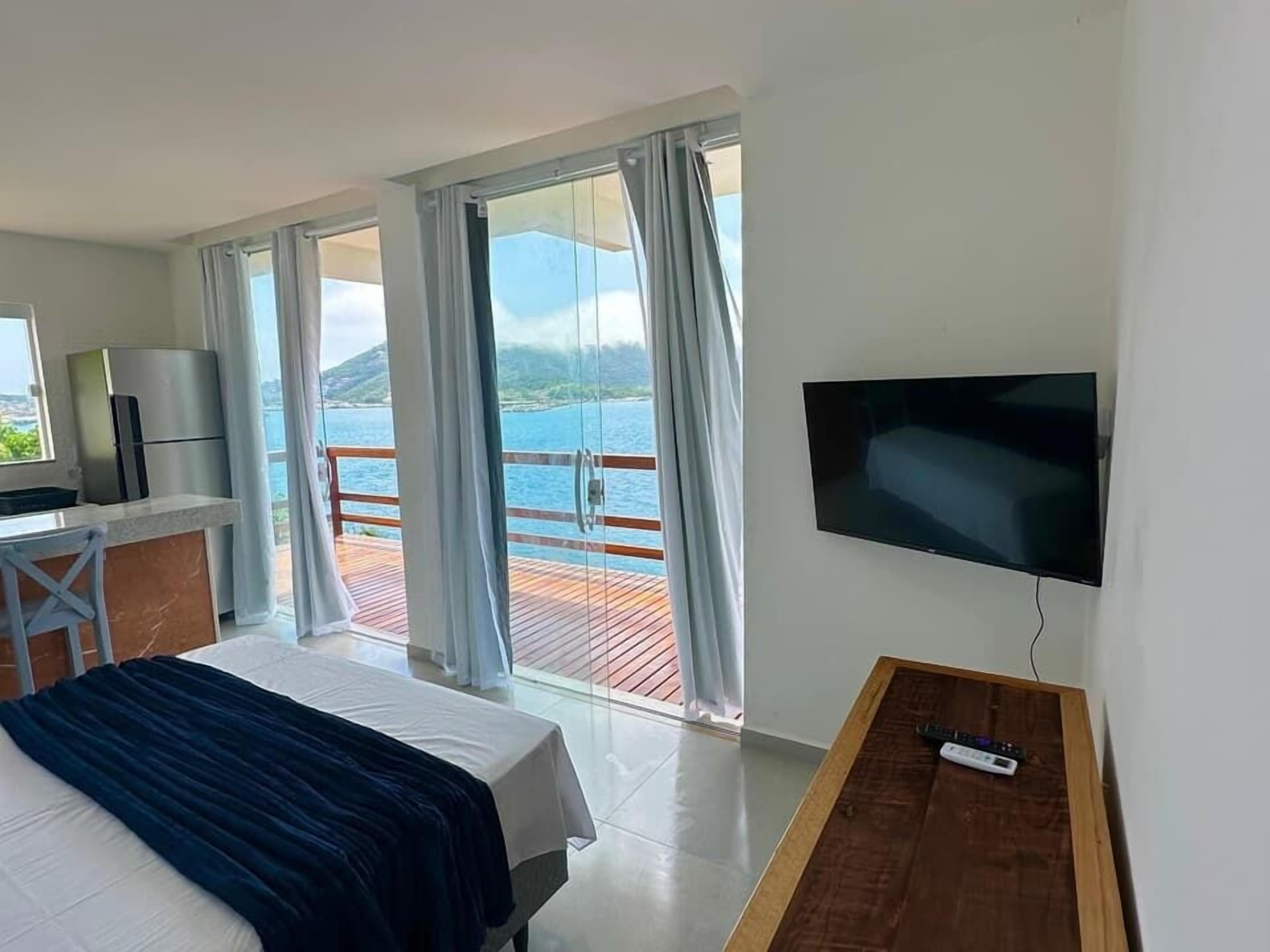 Quarto de hotel com cama, escrivaninha com refrigerador, televisão e varanda vista para o mar.