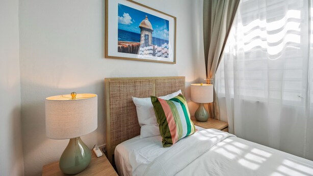 Quarto de hotel bem arrumado, com uma cama, duas luminárias de cabeceira, um quadro na parede e cortinas leves que permitem a entrada de luz natural.