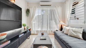 Uma sala de estar moderna com televisão de tela plana, sofá, mesa de centro e uma janela grande com cortinas leves.