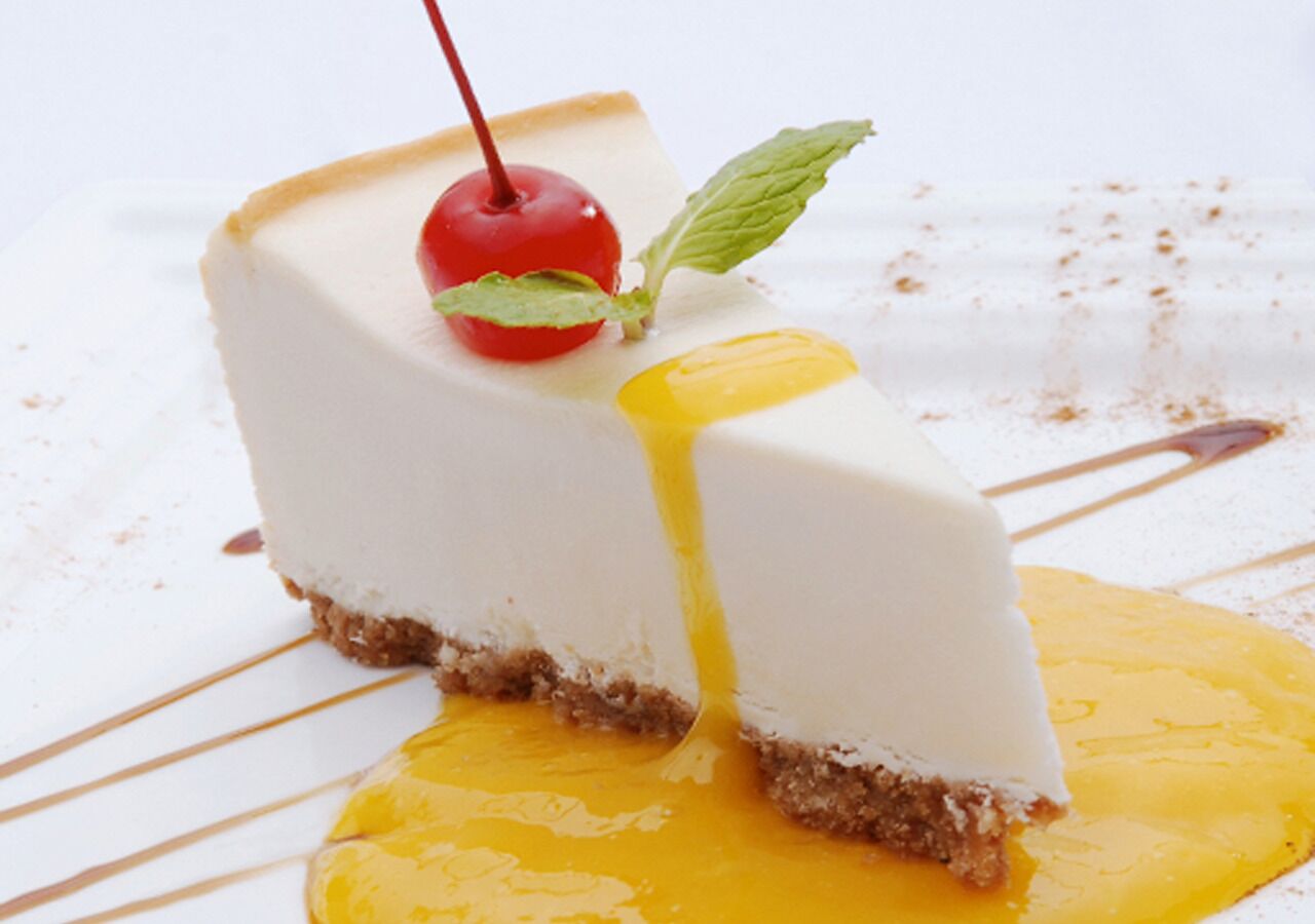 Uma fatia de cheesecake com cereja por cima, uma folha de hortelã e um molho amarelo.