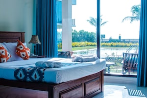 Quarto de hotel com cama grande, vista para piscina e varanda com cadeiras.