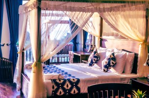 Uma cama de dossel com cortina, roupa de cama branca e almofadas estampadas, um criado-mudo de madeira e vista para o exterior através de uma janela com cortinas azuis.