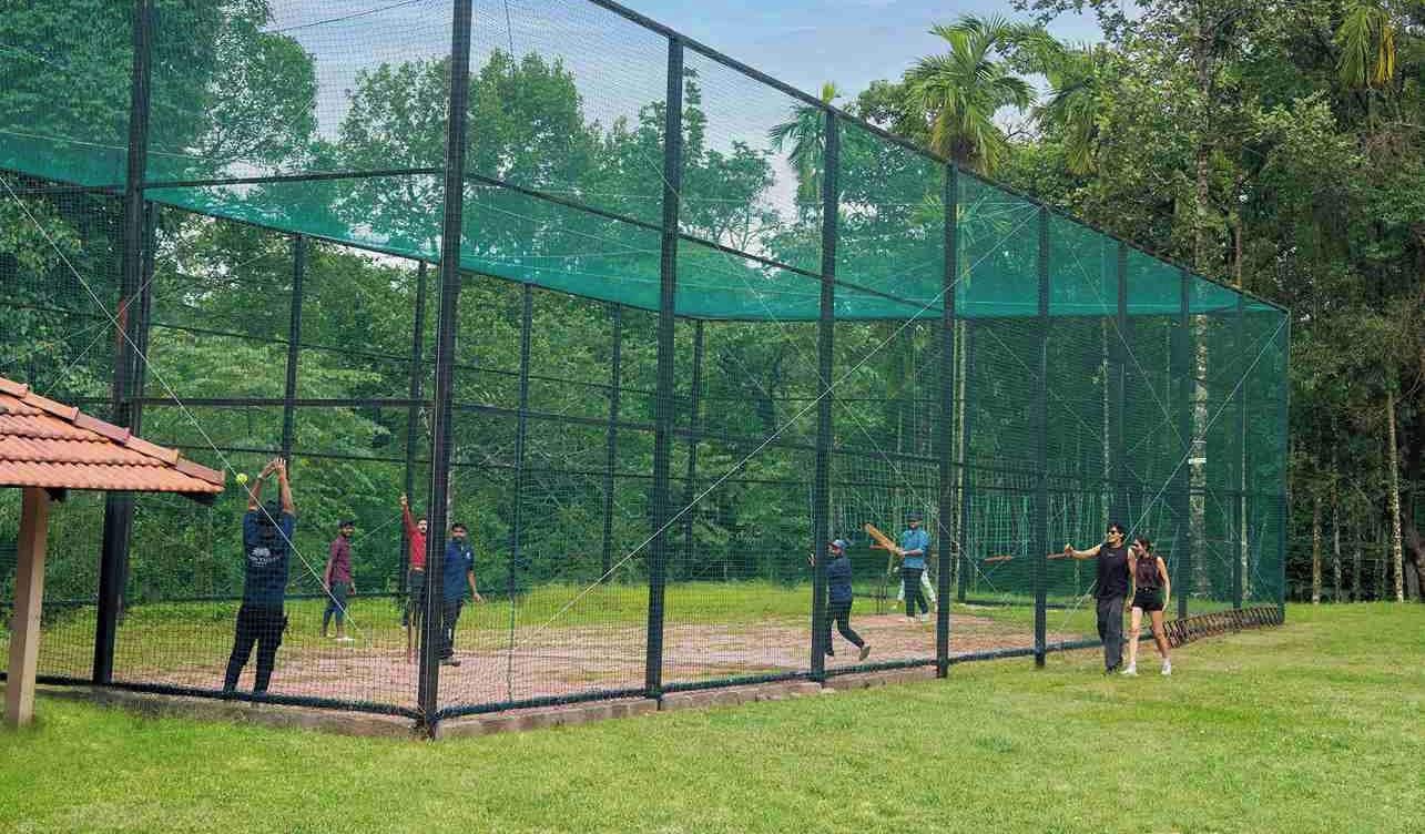Rede de treino de críquete com jogadores em ação, cercada por vegetação exuberante.