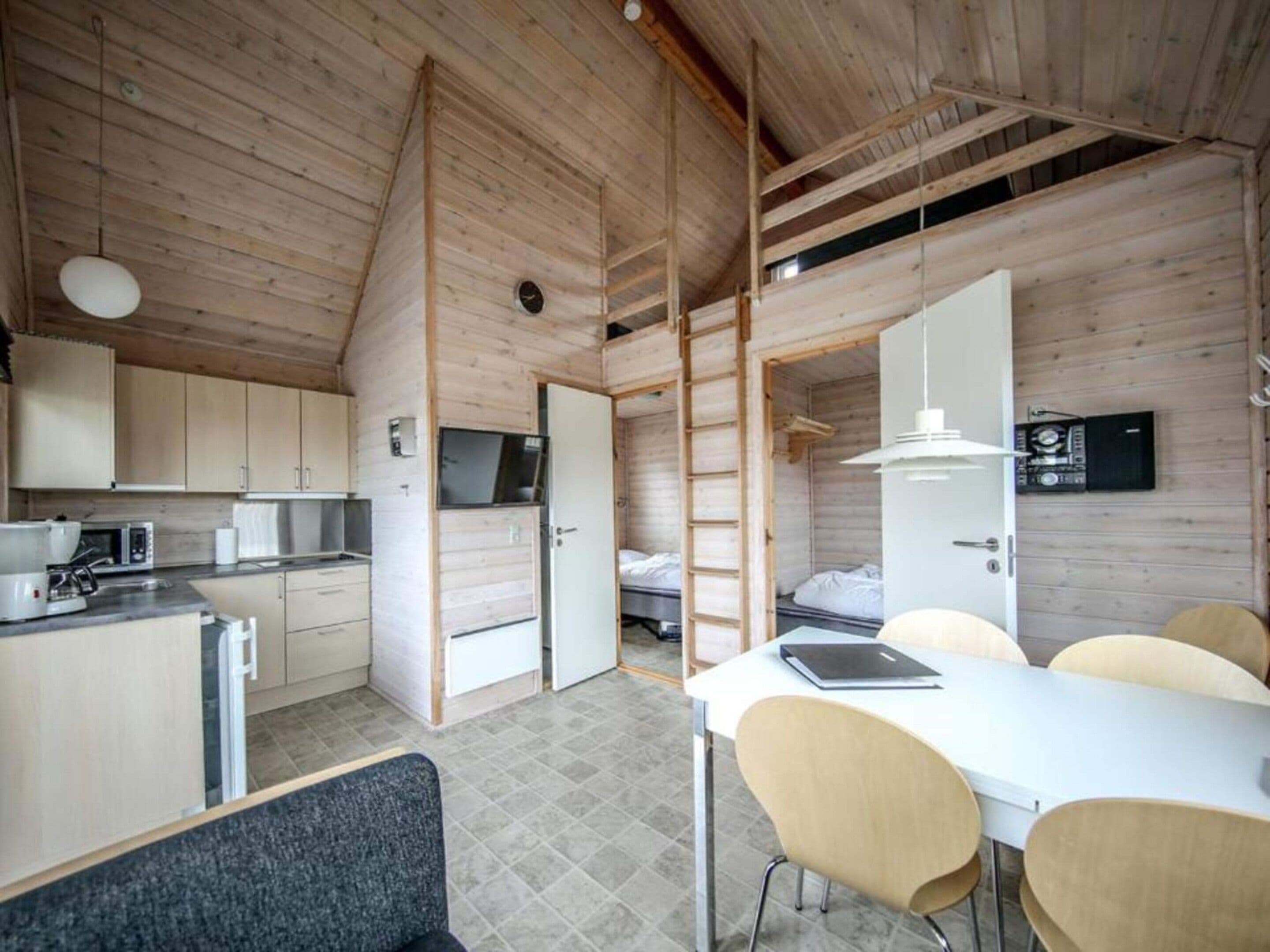 Cozinha compacta e moderna com eletrodomésticos embutidos, área de refeições com mesa e cadeiras, e um mezanino com quarto e escada.