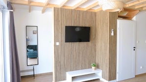 Quarto com televisão presa a uma parede de ripas de madeira, um banco branco, uma porta e um espelho.
