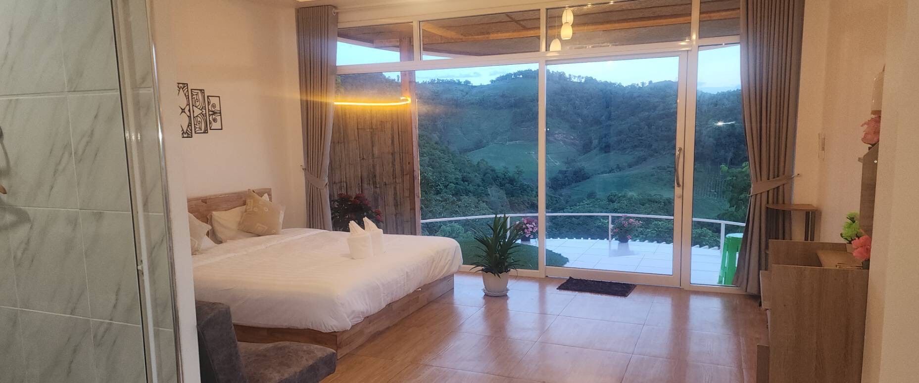 Quarto com uma cama grande, cabeceira de madeira, planta em vaso e varanda com vista para uma paisagem arborizada.