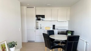 Cozinha moderna com armários brancos, micro-ondas e cafeteira. Há uma mesa redonda com cadeiras pretas e um vaso de planta no balcão.