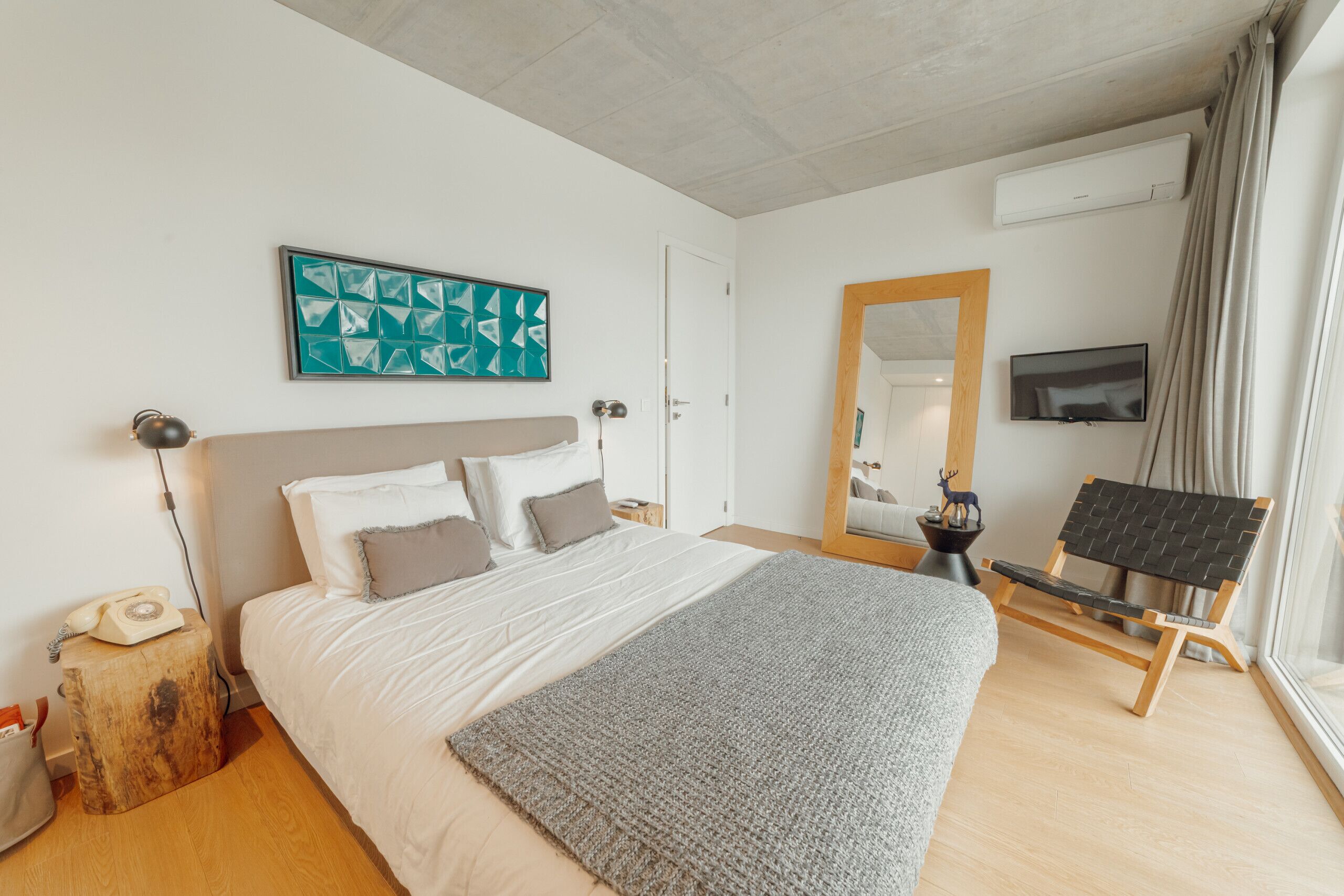 Quarto de hotel moderno com uma cama grande, mesa de cabeceira de madeira, escrivaninha com cadeira, televisão e janela com cortinas.