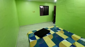 Quarto com paredes verdes, uma cama de solteiro com colcha estampada, uma janela com grades e uma porta.