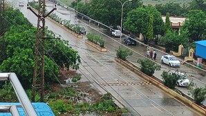 Uma estrada com várias faixas, com veículos, cercada por vegetação e edifícios ao fundo.