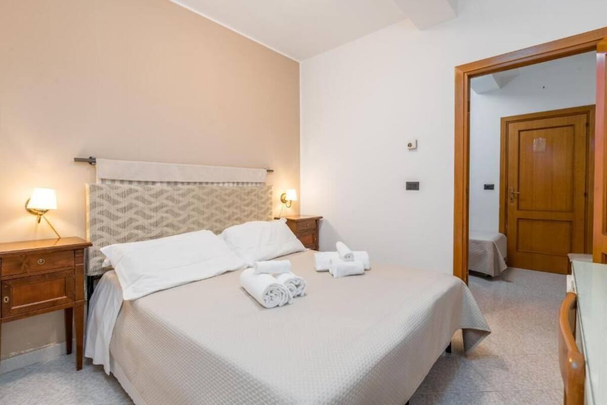 Cama bem arrumada com lençóis e toalhas brancos, uma mesa de cabeceira com abajur e uma porta de madeira ao fundo.