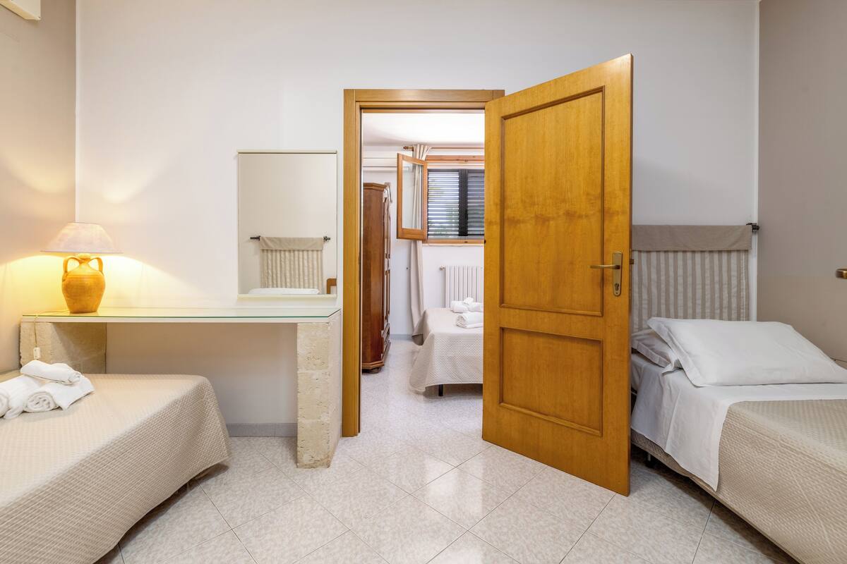 Quarto de hotel com duas camas, mesa de cabeceira com abajur, espelho e porta para o banheiro.