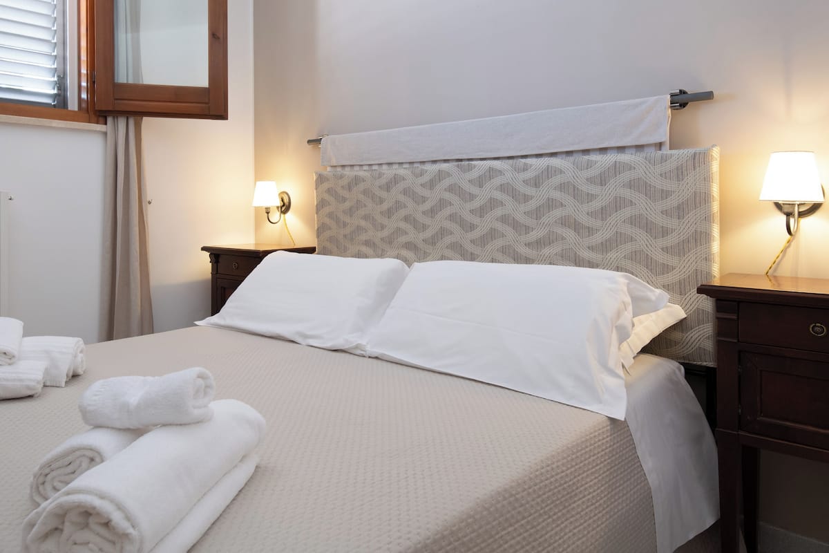 Uma cama bem arrumada com lençóis brancos, duas mesas de cabeceira com abajures e uma janela com persianas.