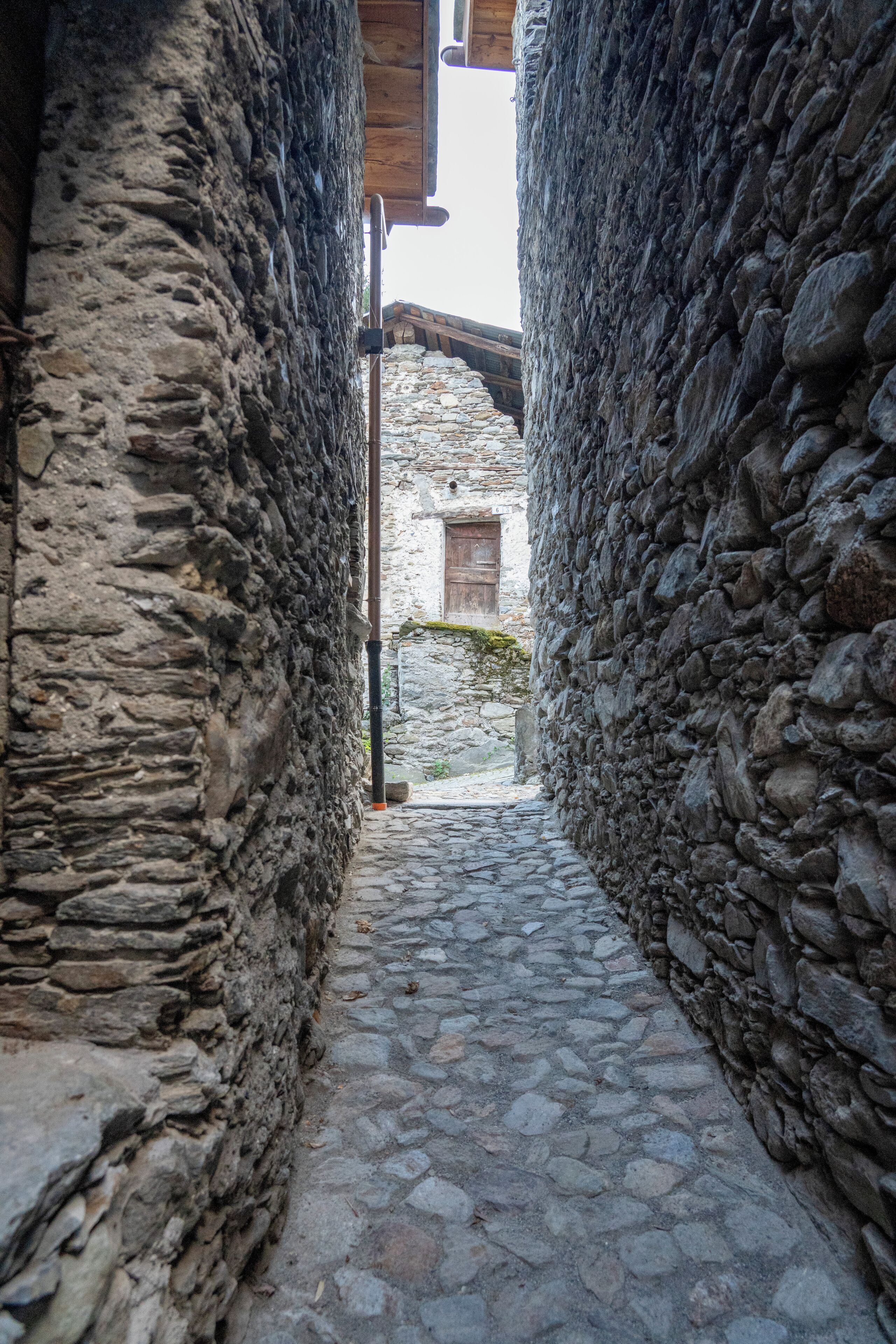 Um caminho de pedra estreito, ladeado por muros de pedra, leva a um edifício rústico com porta de madeira.