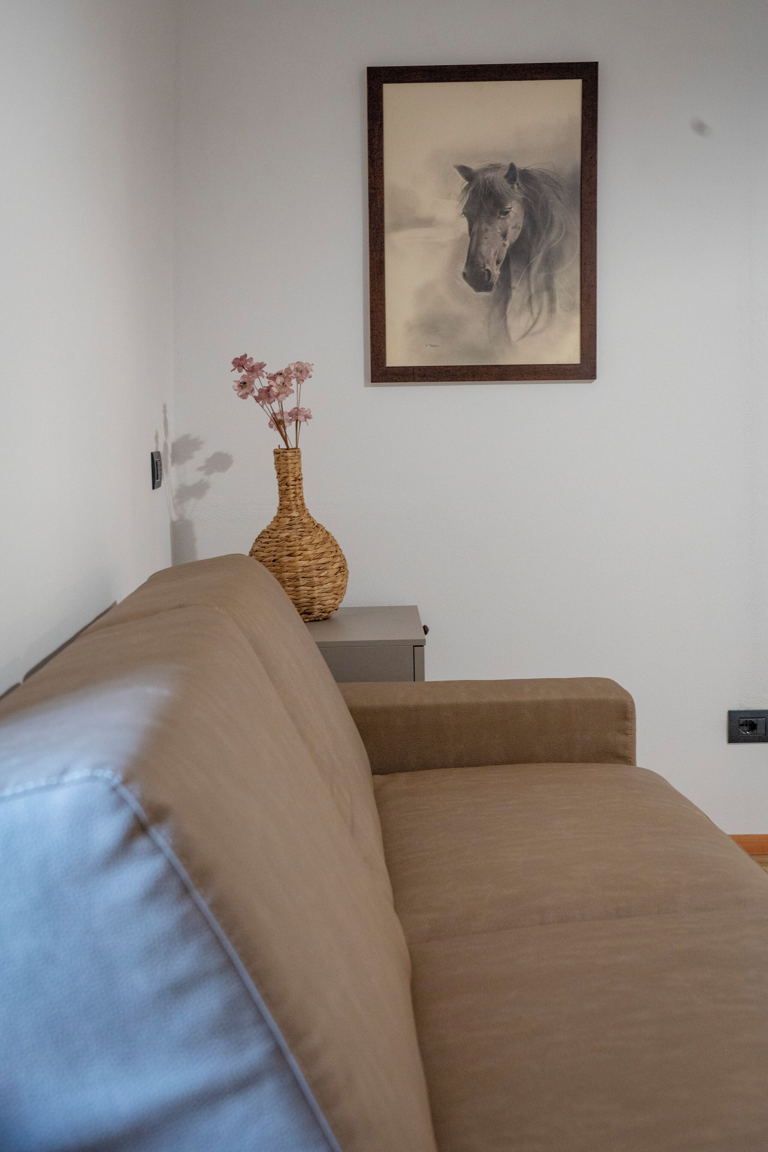 Um quarto com sofá bege, um vaso de vime com flores e um quadro de um cavalo na parede.