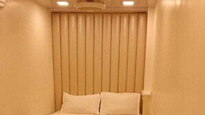 Quarto pequeno e bem arrumado, com uma cama, mesa de cabeceira de madeira e luminária no teto.