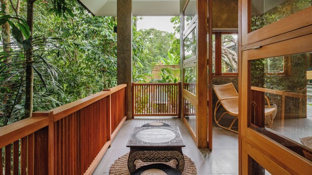 Varanda de madeira com vista para vegetação exuberante, uma cadeira de fibra e uma mesa pequena com tampo decorativo.