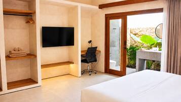 Quarto de hotel com cama, escrivaninha e cadeira, televisão, guarda-roupa e banheiro visível por uma porta de vidro.